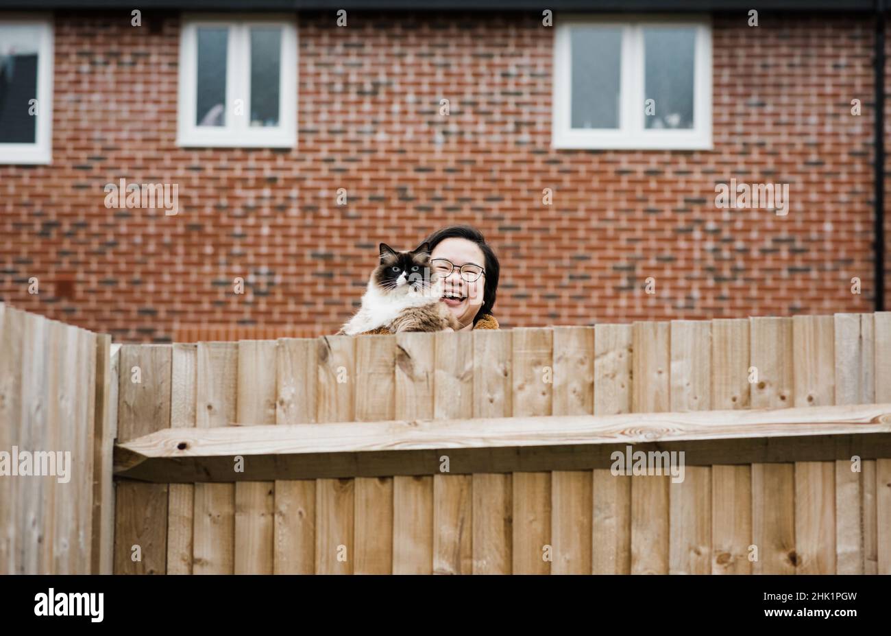 Woman with her cat smiling over garden fence saying hello to neighbour ...