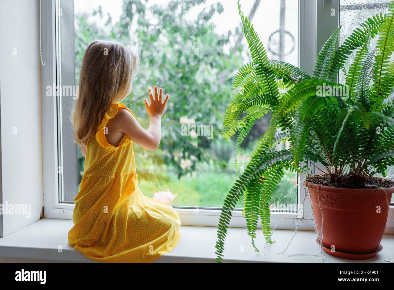 Young little blonde girl backside sitting on window sill thoughtfully ...