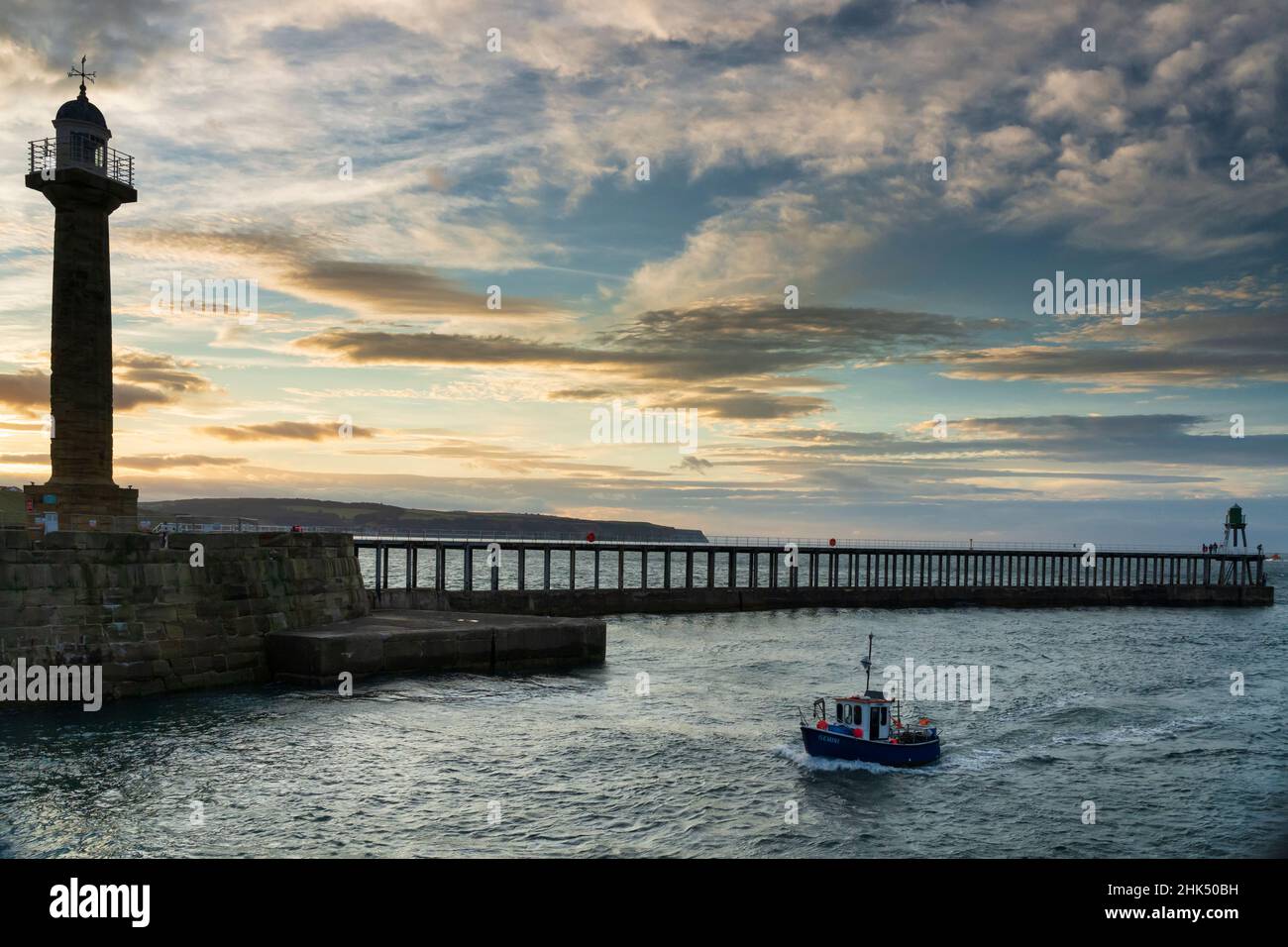Sunset over Whitby harbour, pier and lighthouse as a small fishing boat ...