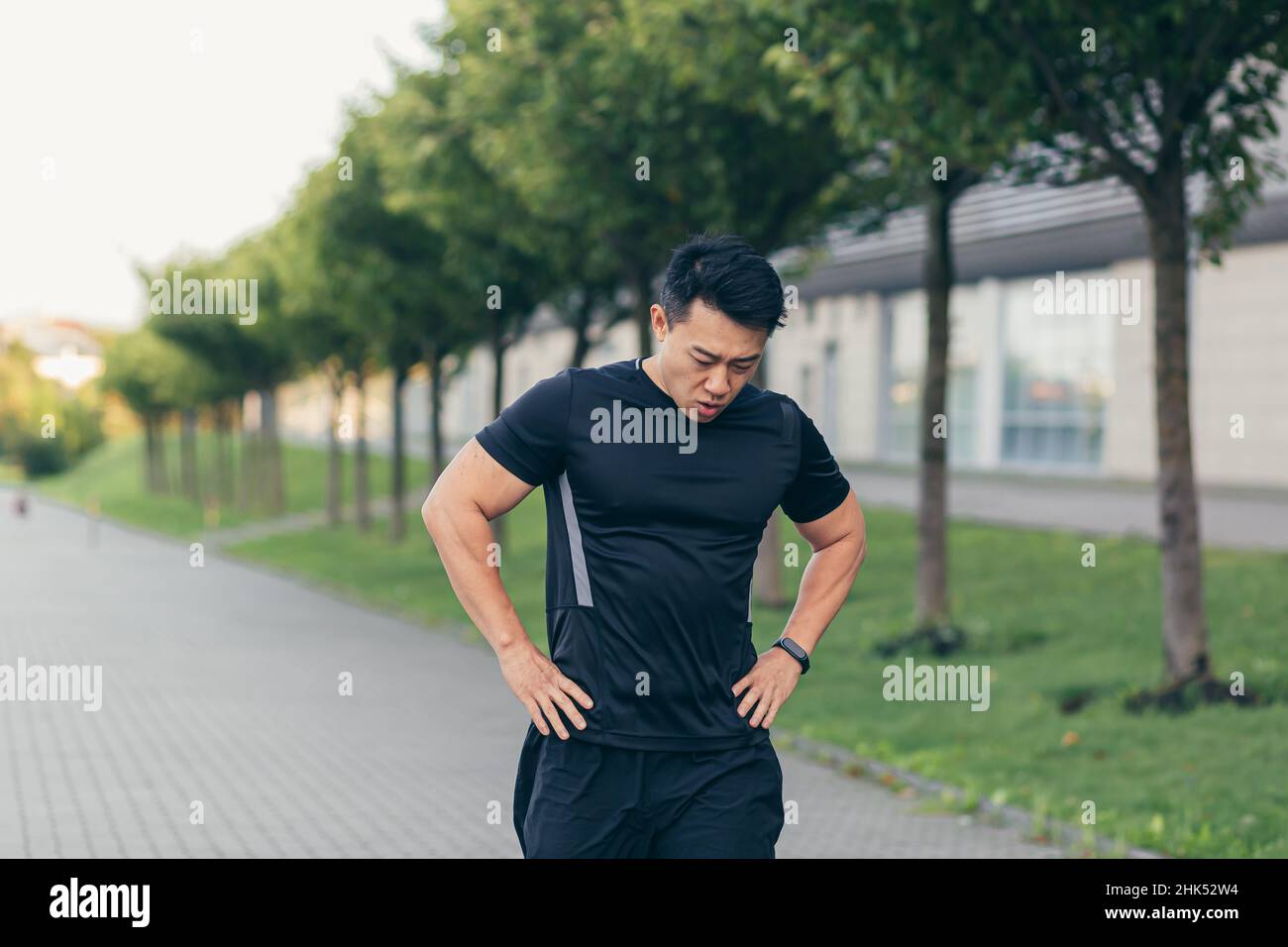 Male asian athlete, tired after a morning jog, runs in the park near ...