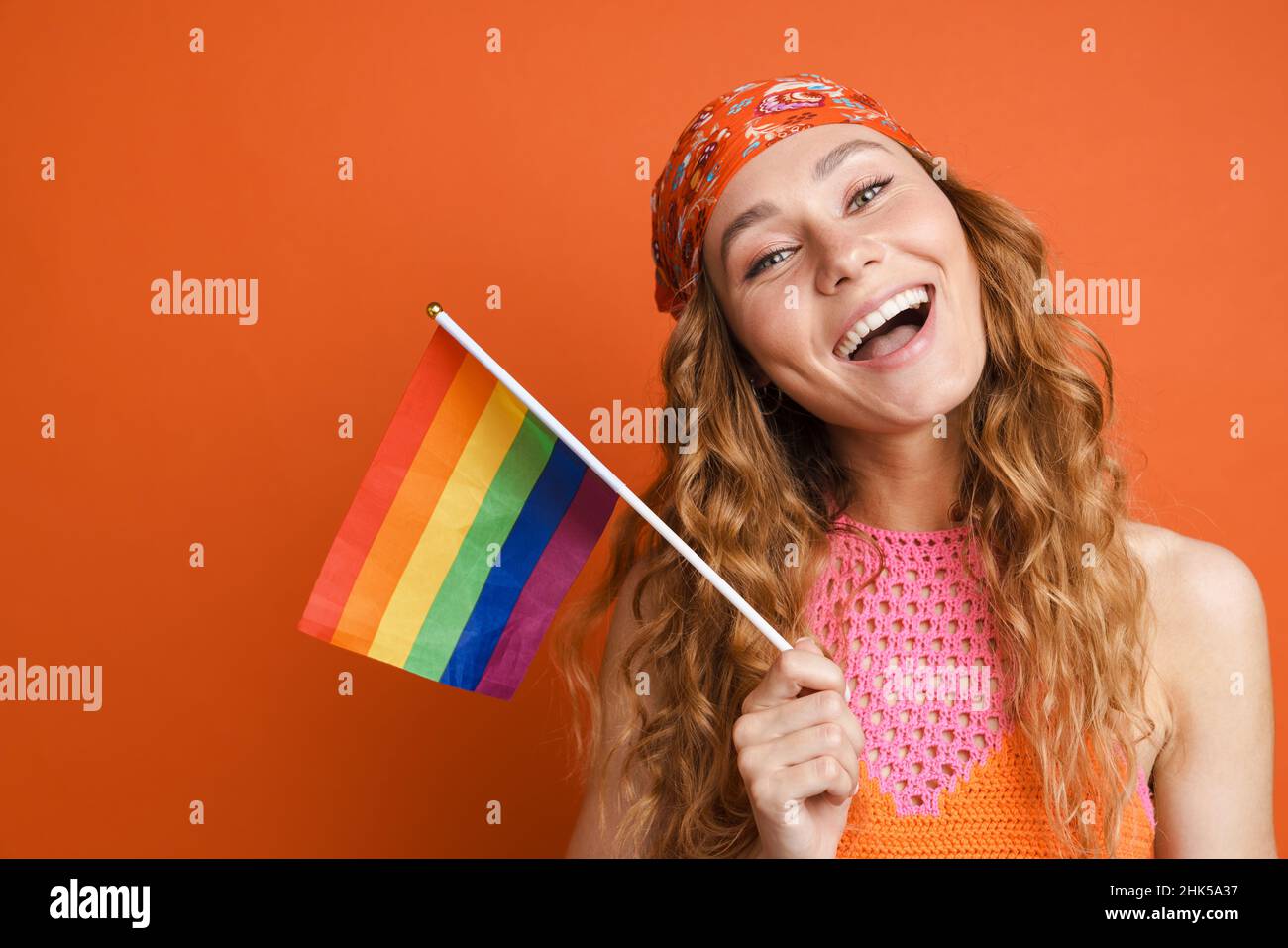 Young ginger woman in bandana smiling while posing with rainbow flag ...