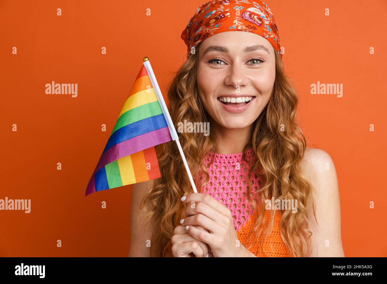 Young ginger woman in bandana smiling while posing with rainbow flag ...