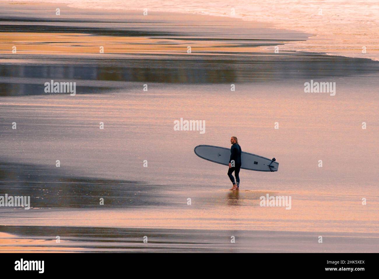 Warm evening light over a surfer carrying his surfboard and walking on ...