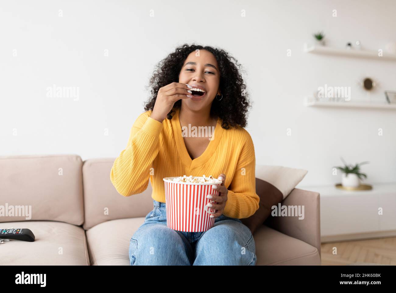 Lovely young black woman eating popcorn from bucket while watching TV ...