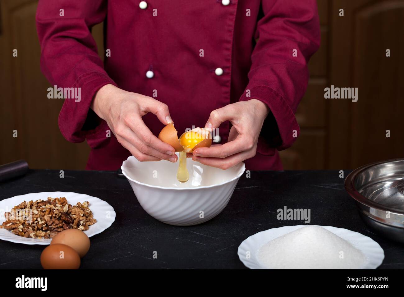 A professional chef in a burgundy jacket breaks an egg into a glass ...