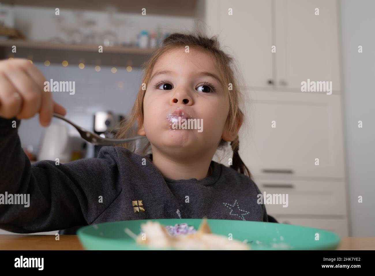 Little child girl sitting at the table and putting spoon at her mouth ...