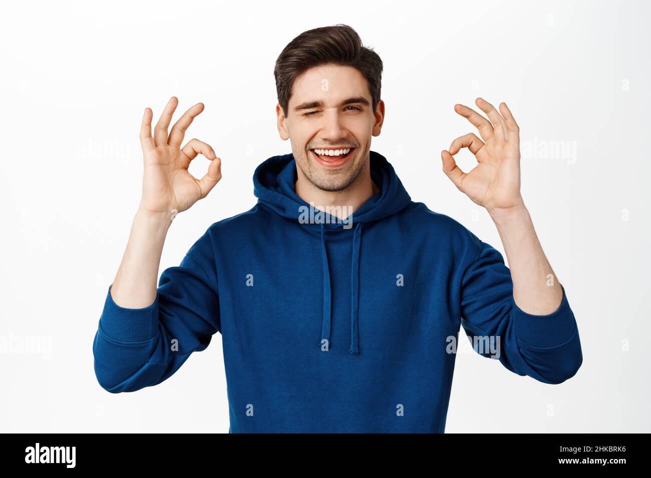 Young millennial man winks and smiles satisfied, shows okay, ok signs ...