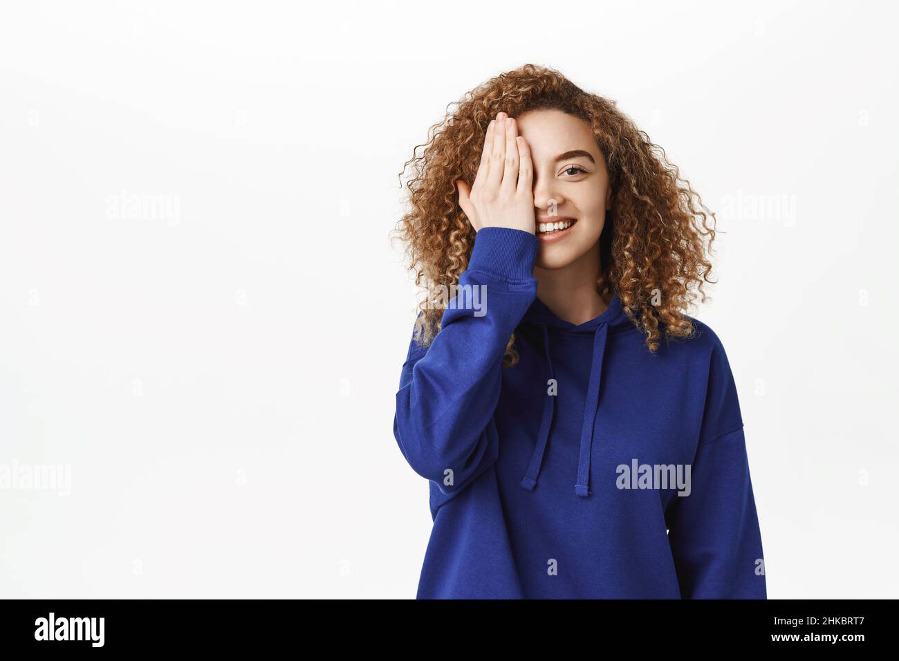 Smiling girl covers one side of face, looking with half, standing in ...