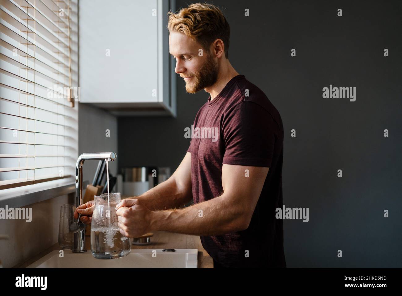 Smiling man pouring water in the jug from a tap while standing at the ...