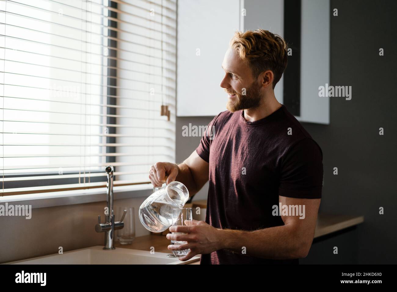 Attractive young man pouring water from a juf in a glass while standing ...
