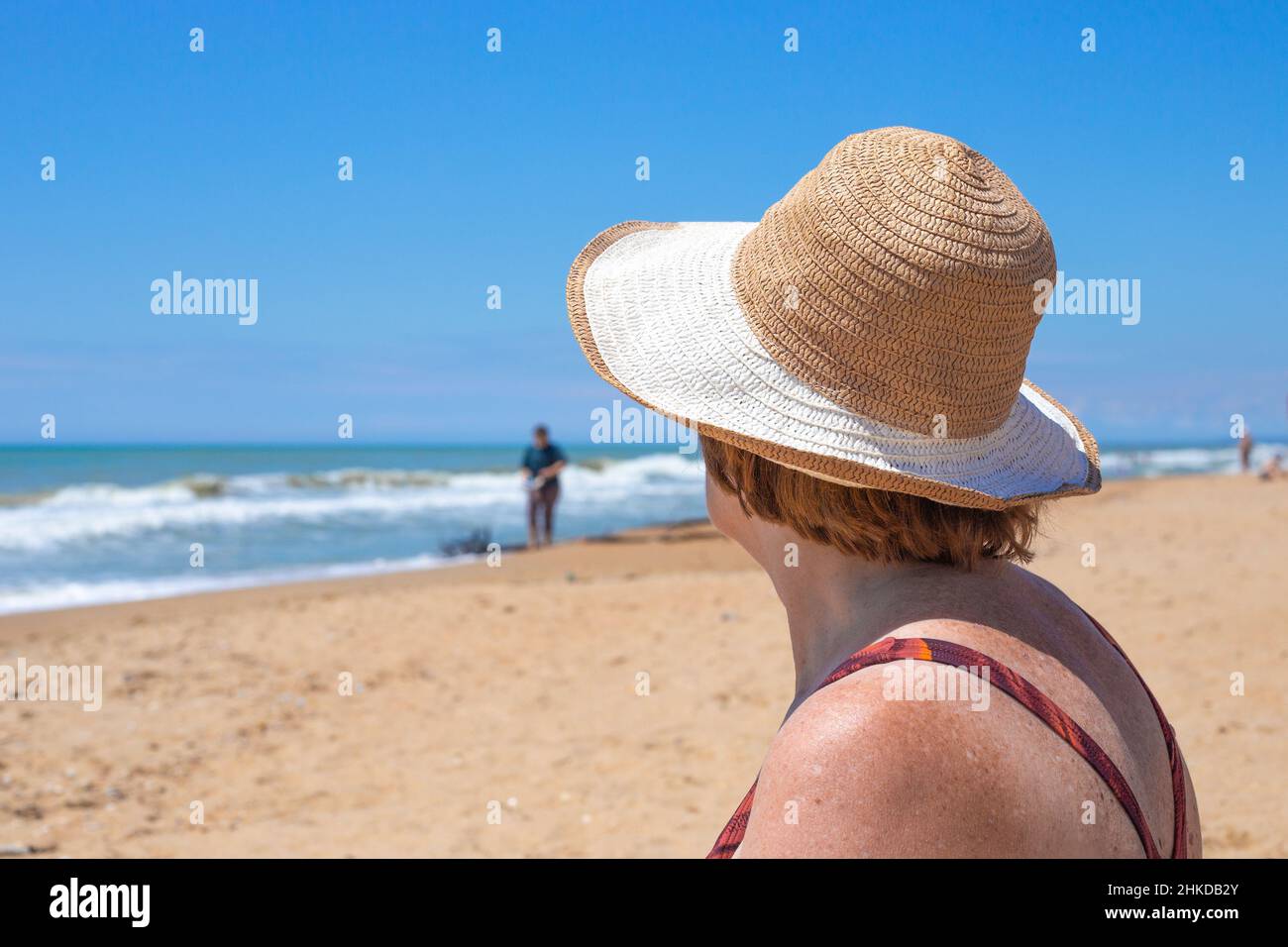 Mature woman in a hat on the sandy shore looks at the sea, side view ...