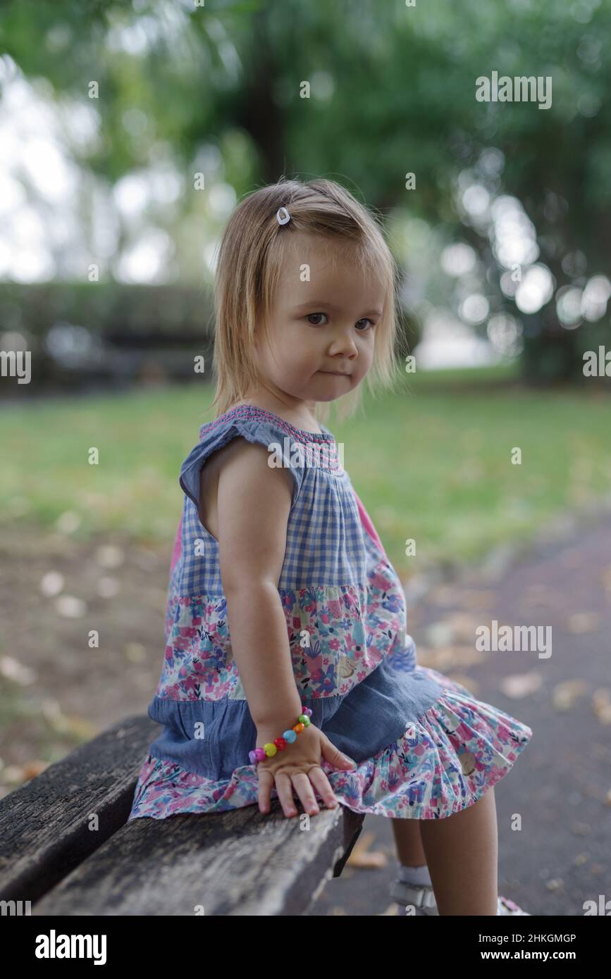 Lovely toddler girl sitting on bench in the park while looking away ...