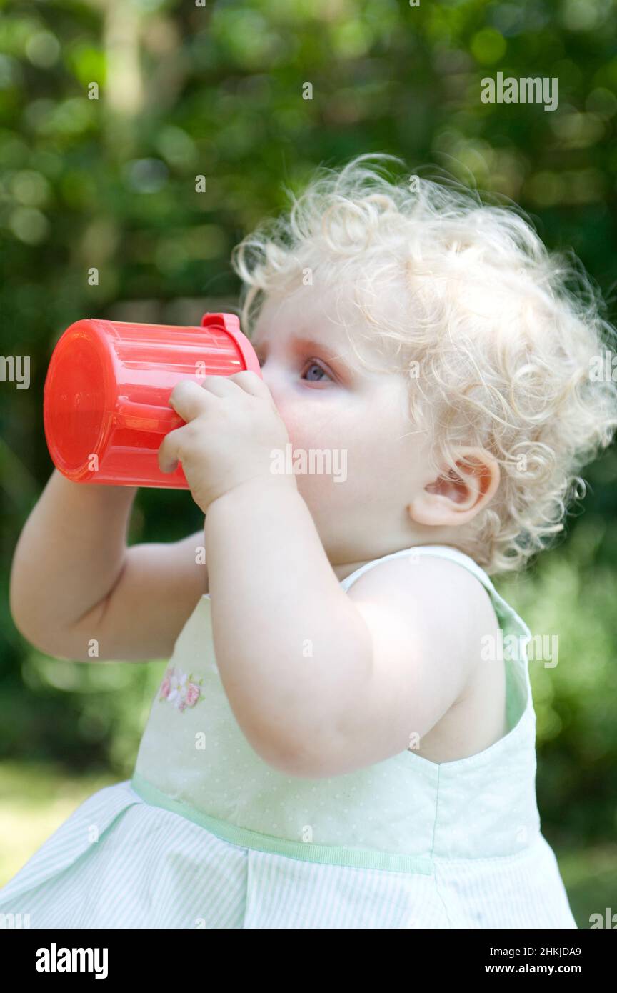 Baby girl in a garden drinking from red plastic beaker Stock Photo - Alamy