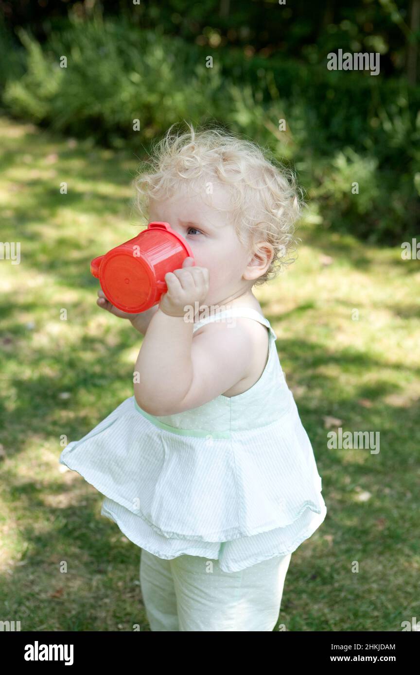 Baby girl in a garden drinking from red plastic beaker Stock Photo - Alamy