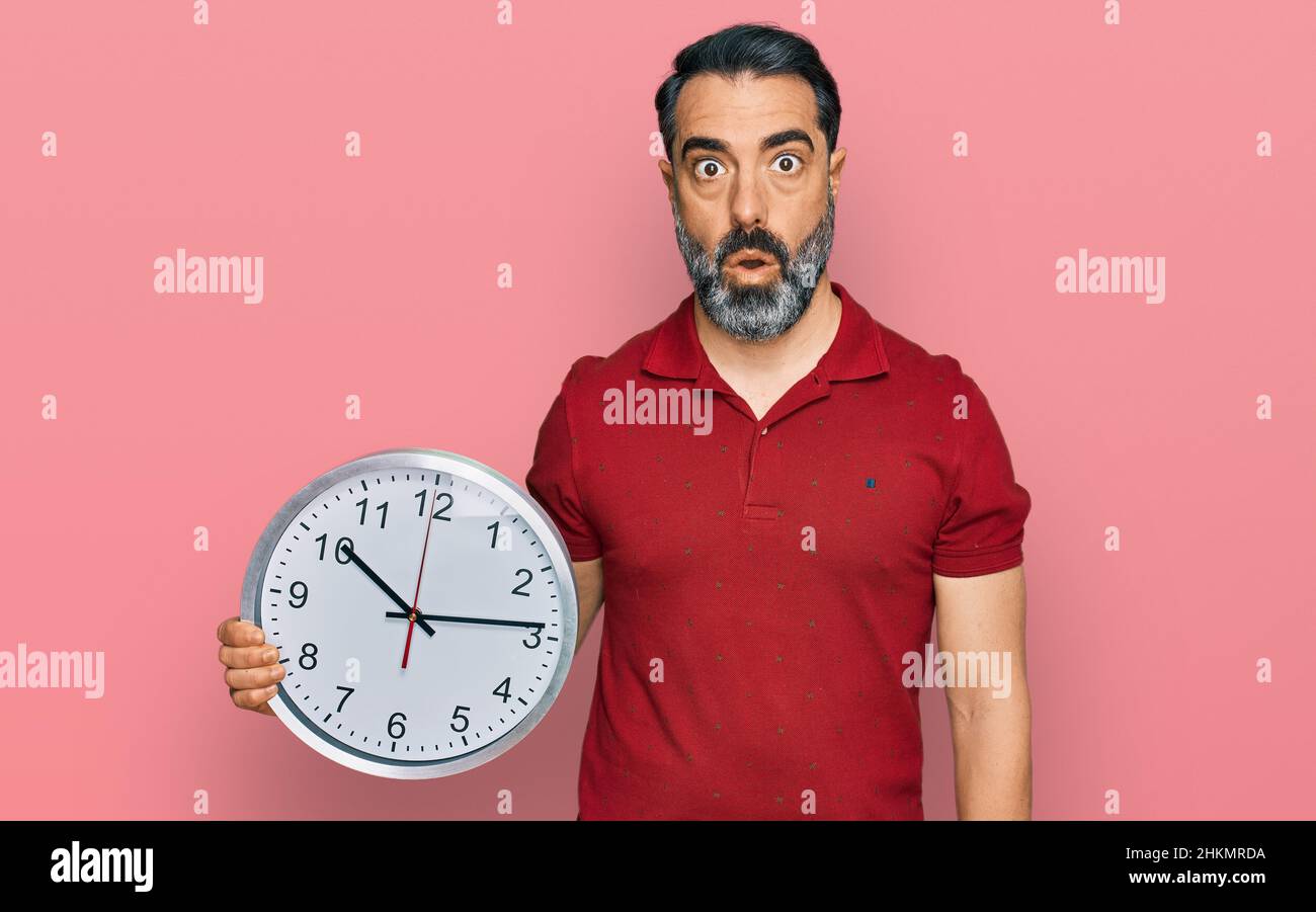 Middle aged man with beard holding big clock scared and amazed with ...