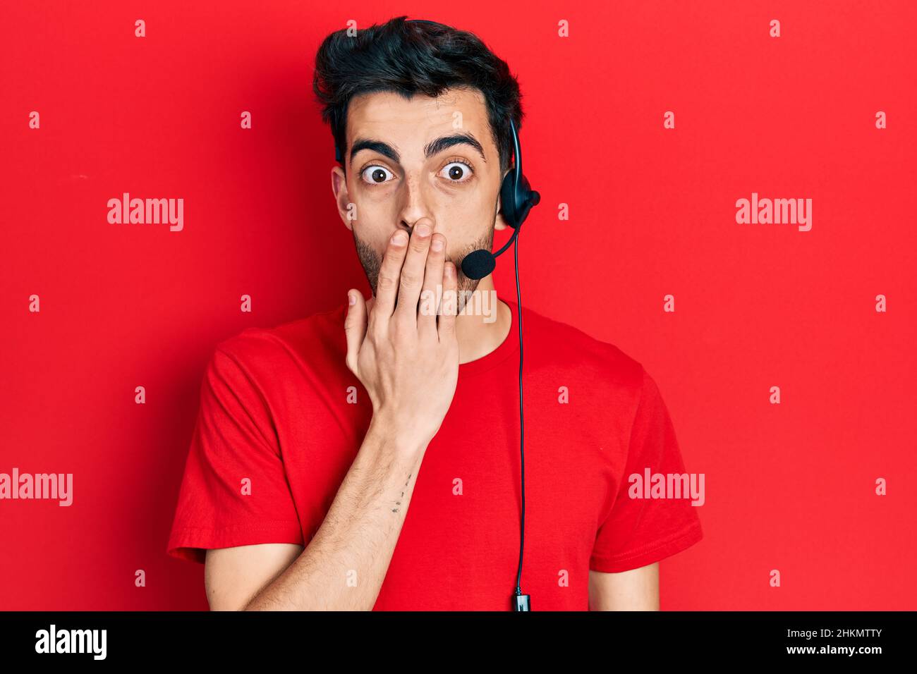 Young hispanic man wearing call center agent headset covering mouth ...