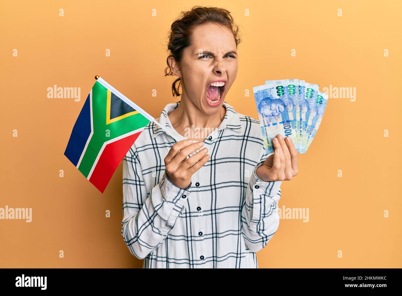 Young brunette woman holding south africa flag and rand banknotes angry ...