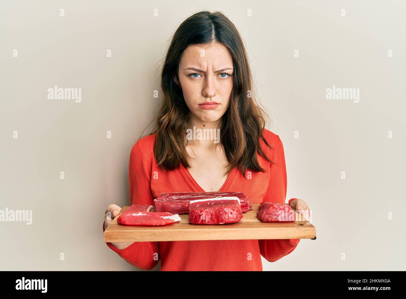 Young brunette woman holding board with raw meat depressed and worry ...