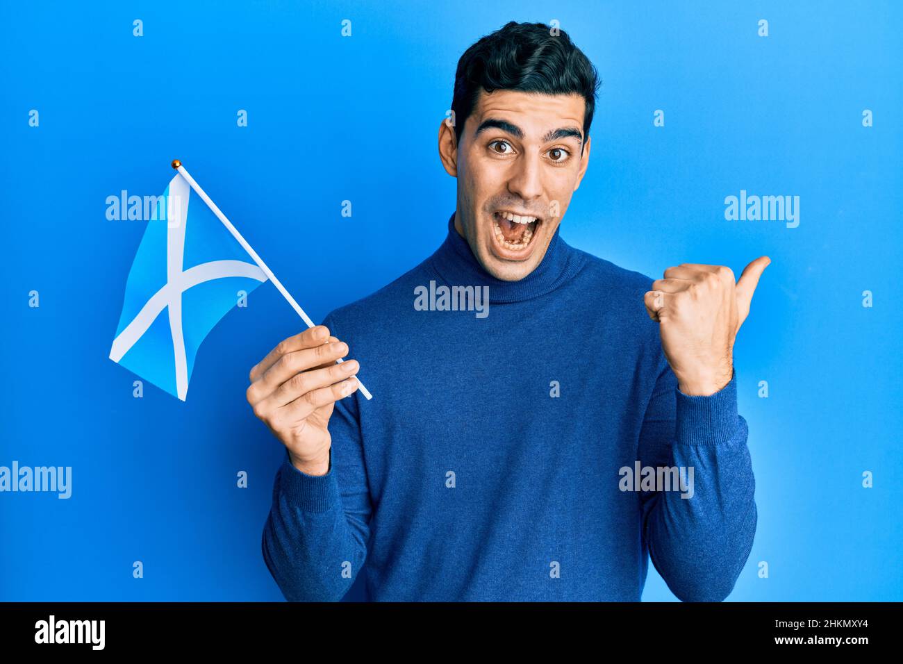 Handsome hispanic man holding scotland flag pointing thumb up to the ...