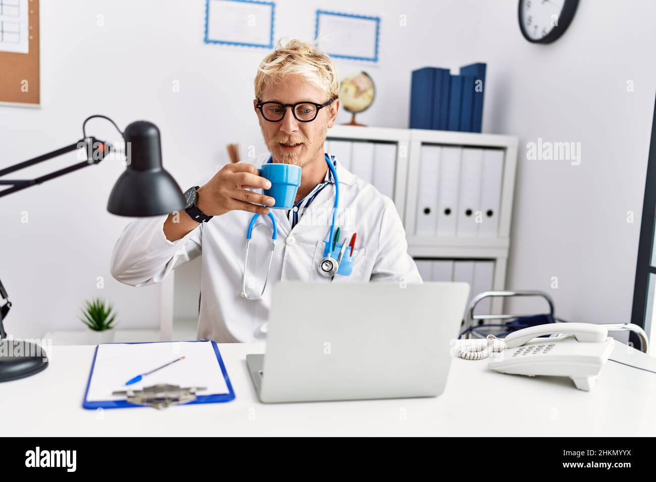 Young caucasian man wearing doctor uniform drinking coffee at clinic ...