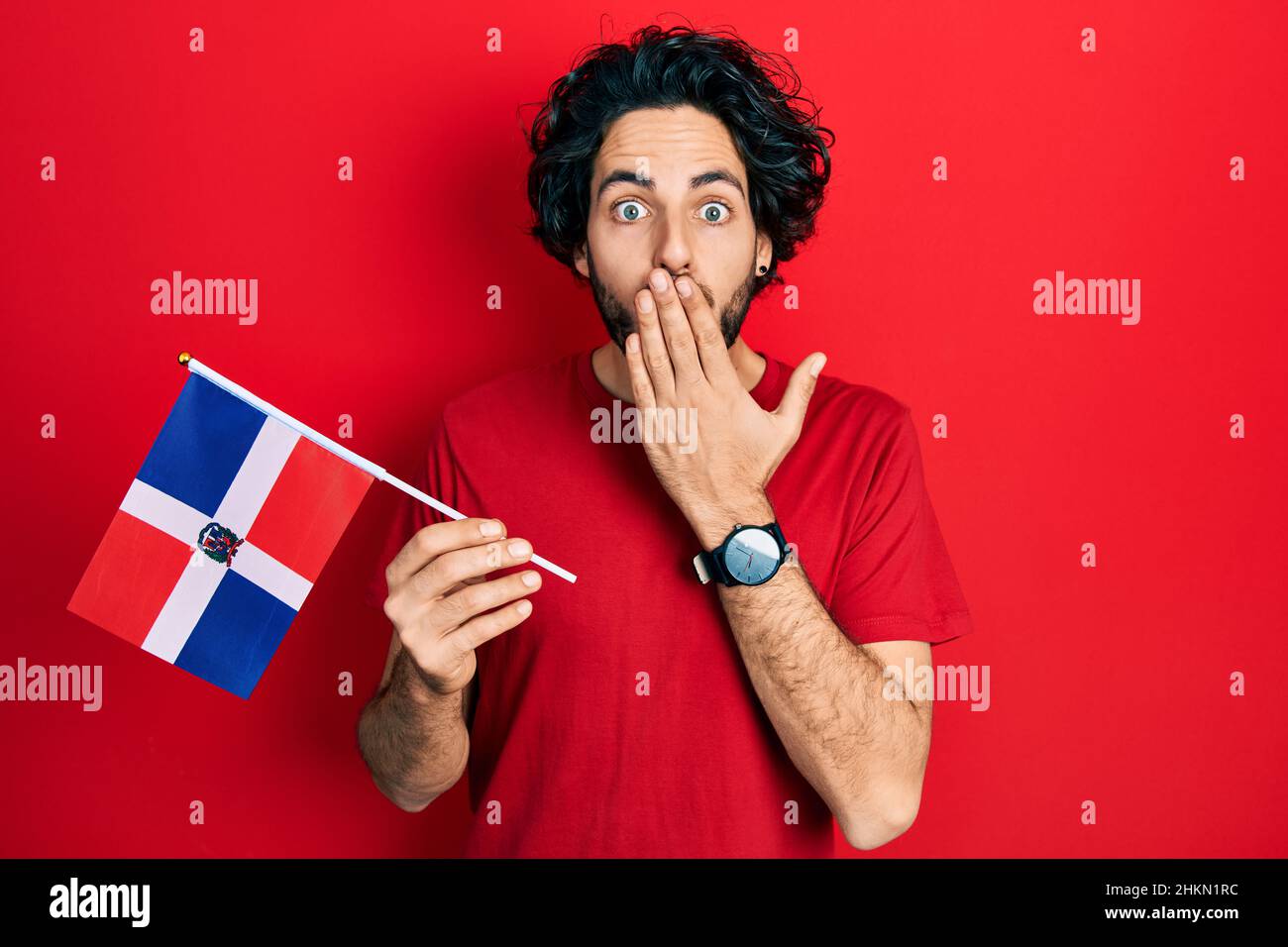 Handsome hispanic man holding dominican republic flag covering mouth ...