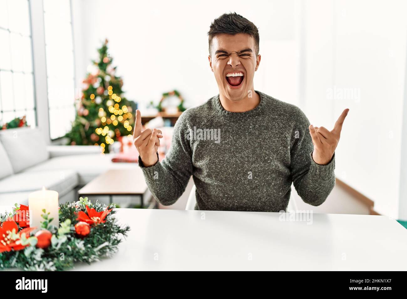 Young hispanic man sitting on the table by christmas tree shouting with ...