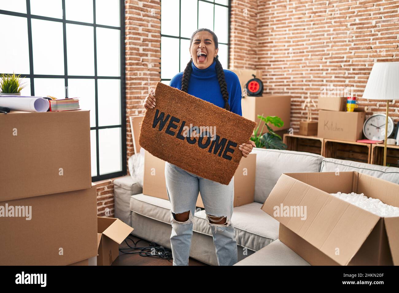 Young hispanic woman holding welcome doormat at new home angry and mad ...