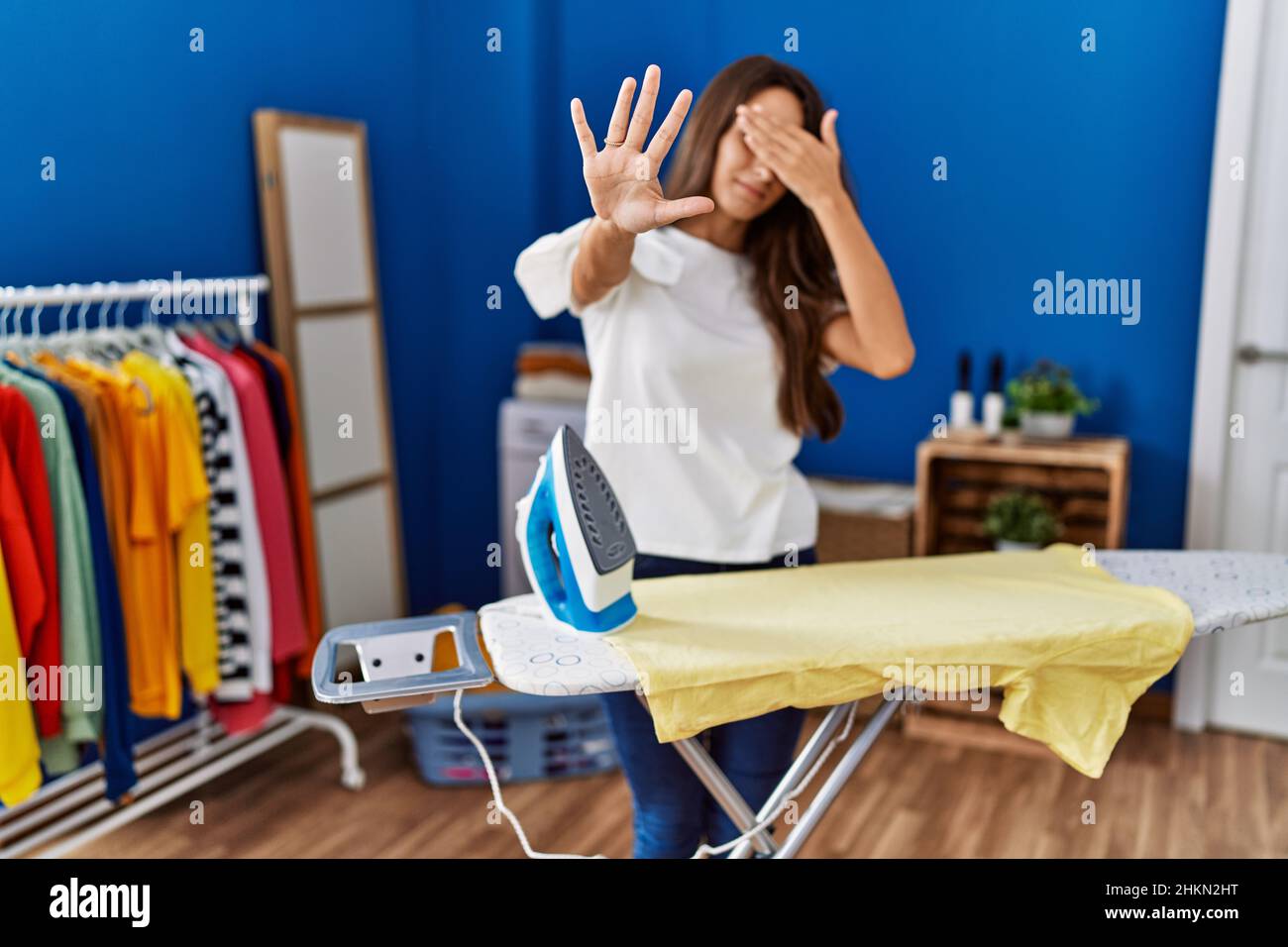 Young hispanic woman ironing clothes at laundry room covering eyes with ...