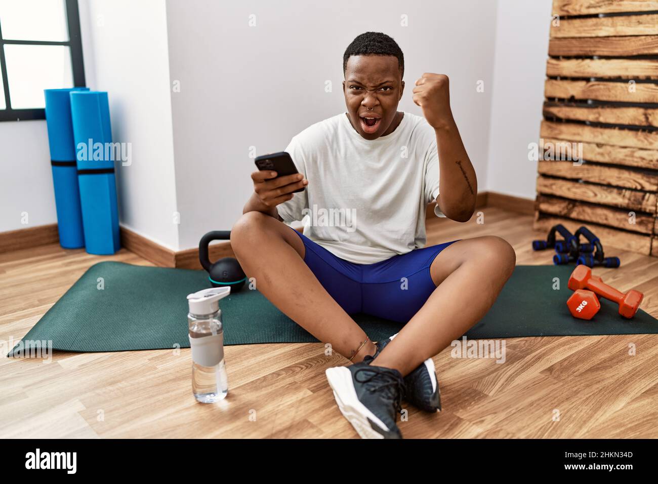 Young african man sitting on training mat at the gym using smartphone ...