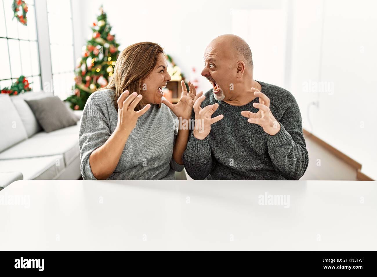 Middle age hispanic couple sitting on the table by christmas tree crazy ...