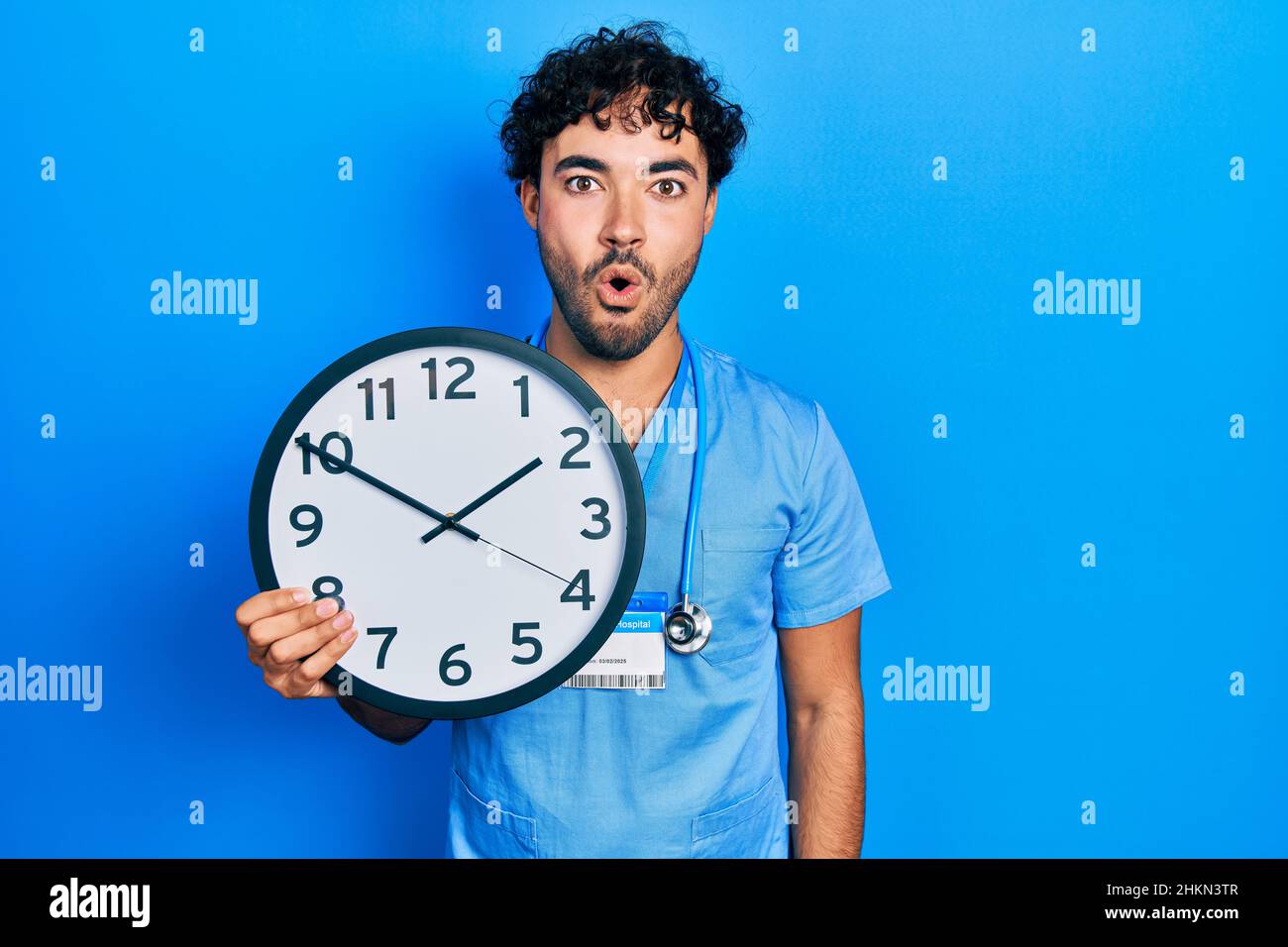 Young hispanic man wearing blue male nurse uniform holding clock scared ...