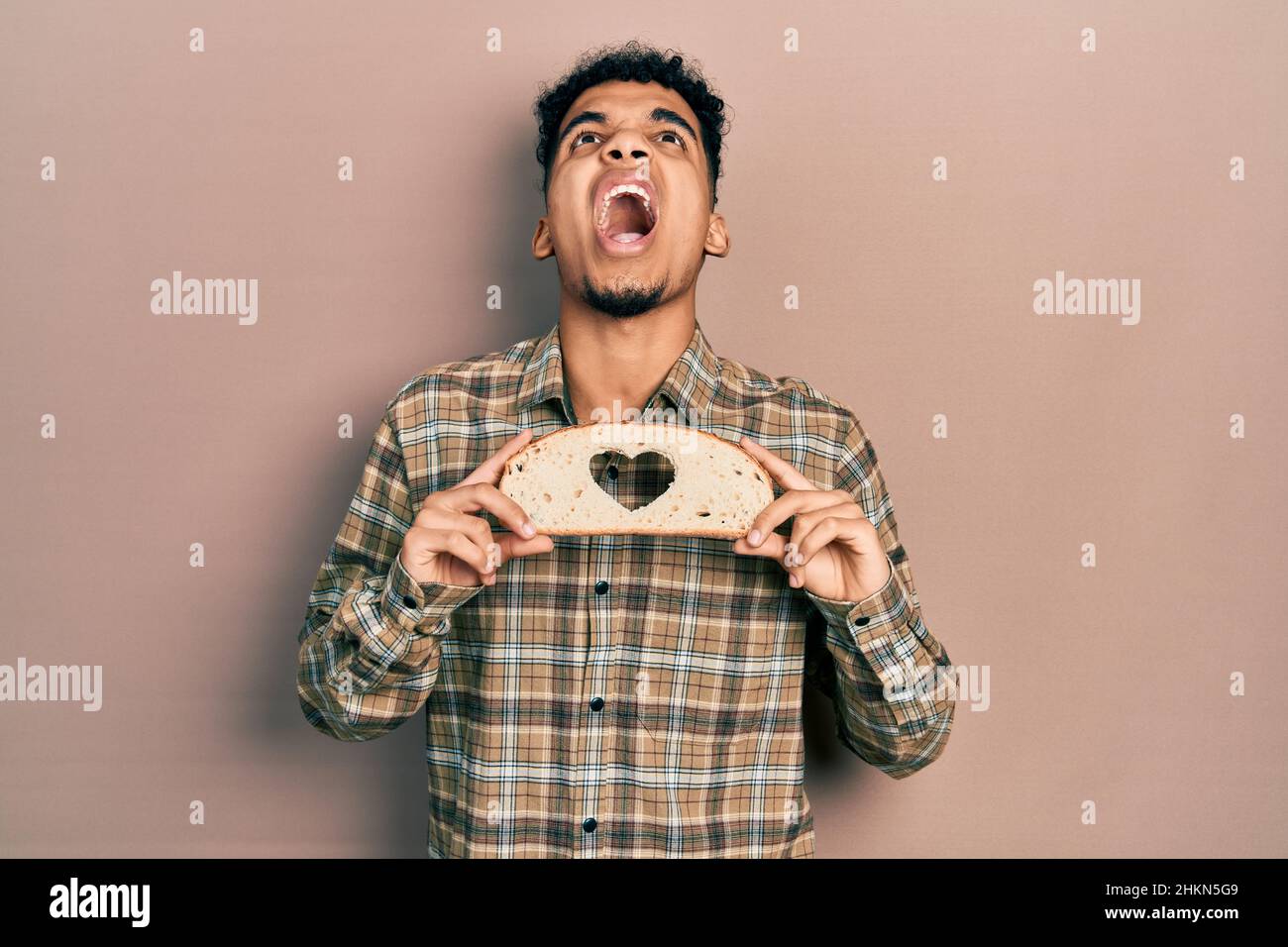 Young african american man holding bread loaf with heart shape angry ...