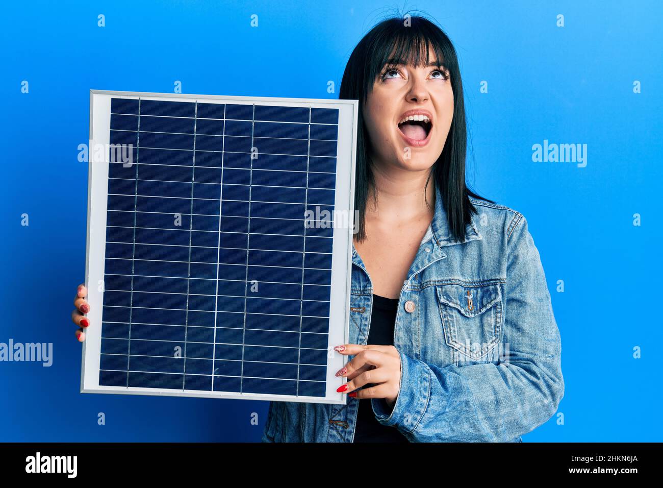 Young hispanic woman holding photovoltaic solar panel angry and mad ...