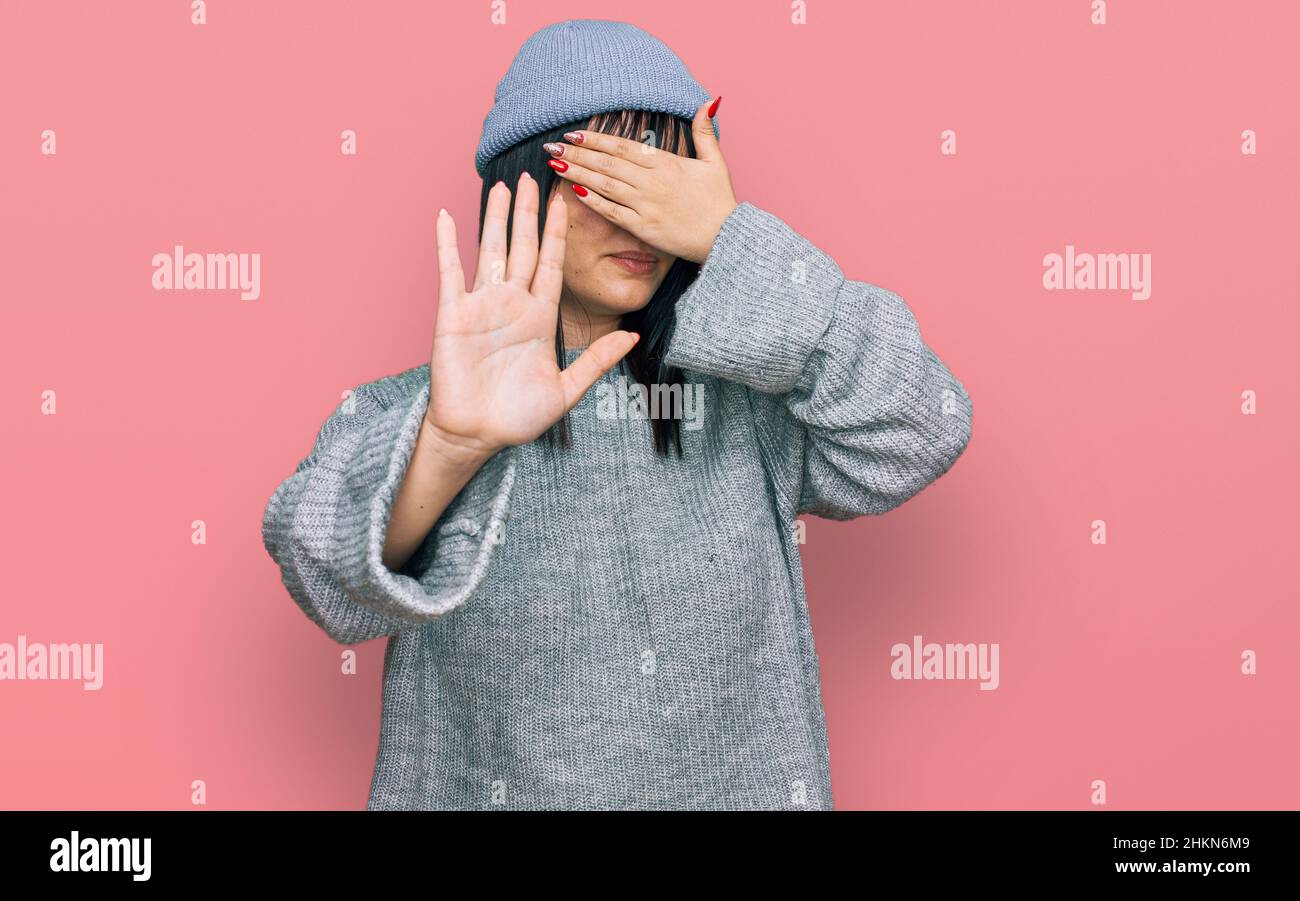 Young hispanic woman wearing cute wool cap covering eyes with hands and ...