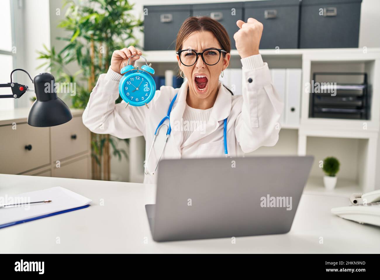 Young brunette doctor woman holding alarm clock annoyed and frustrated ...