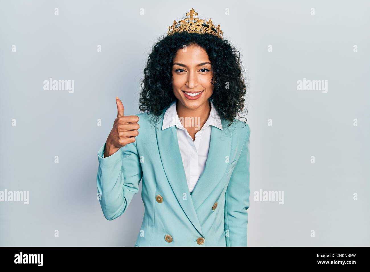Young latin girl wearing business clothes and queen crown smiling happy and positive, thumb up ...