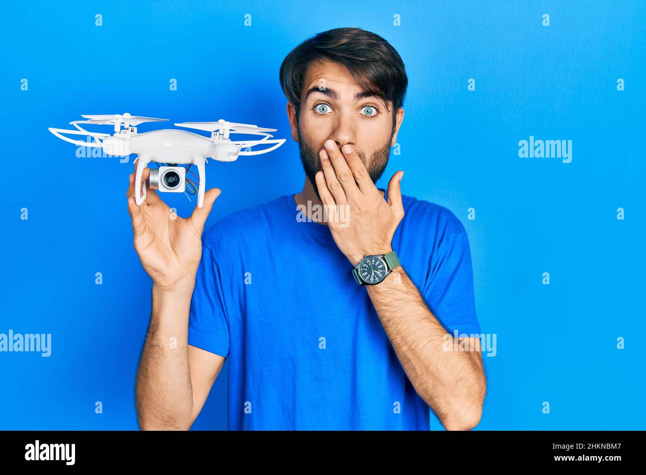 Young hispanic man holding drone covering mouth with hand, shocked and ...
