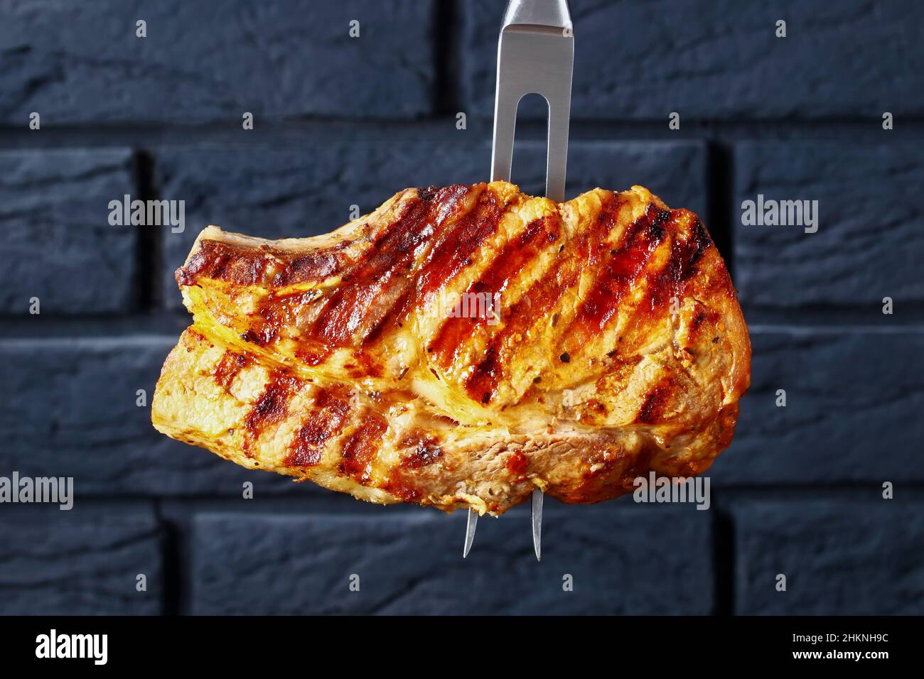 carving fork with speared grilled pork chop on it with a black brick ...