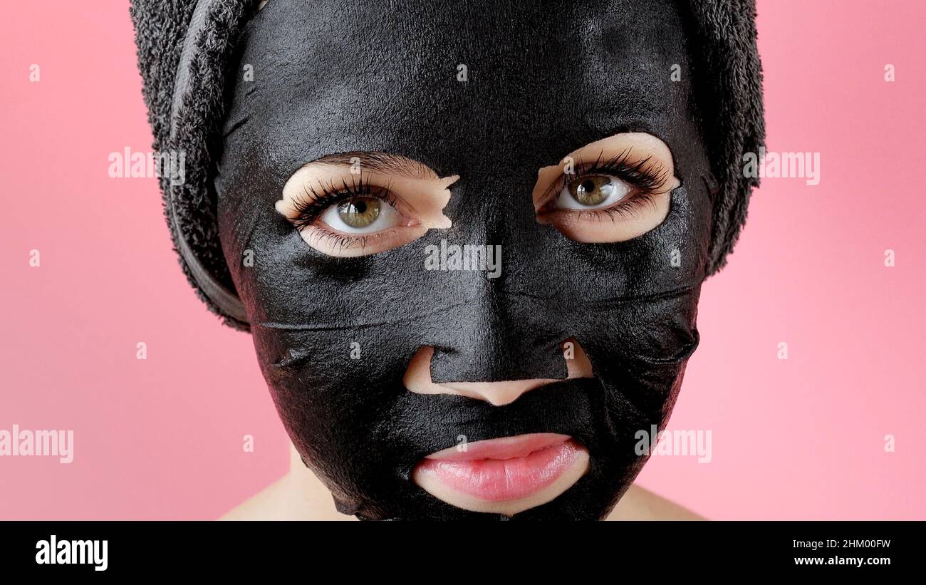 Young woman apply black cosmetic fabric facial mask on pink background ...