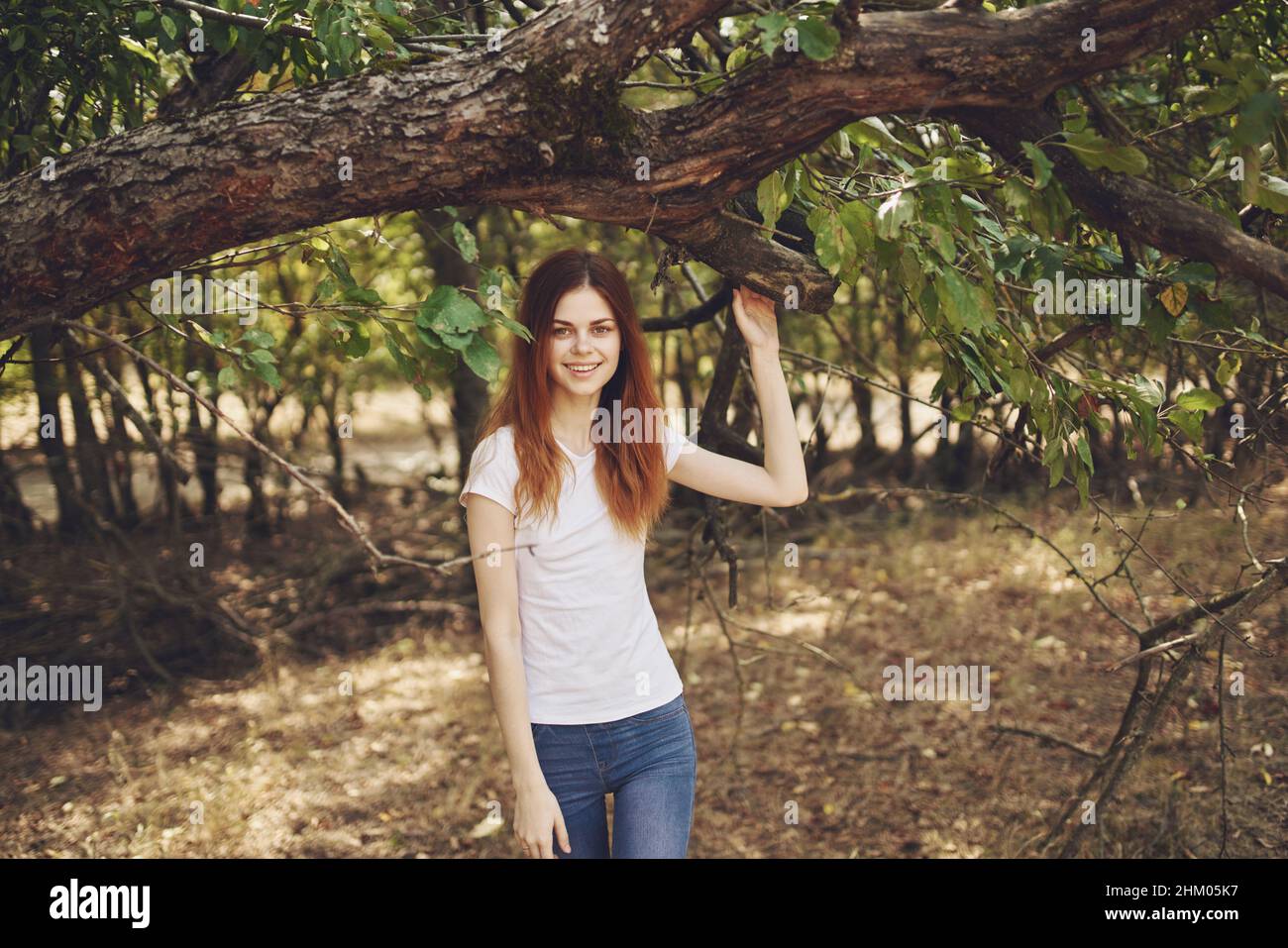 woman in a t-shirt and jeans stands near a tree with green leaves on ...