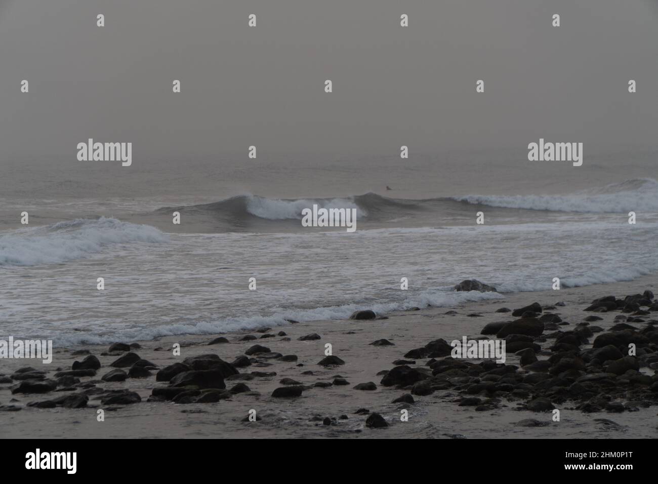 Distant view of the ocean, waves, rocks and the surfer on a gloomy day ...