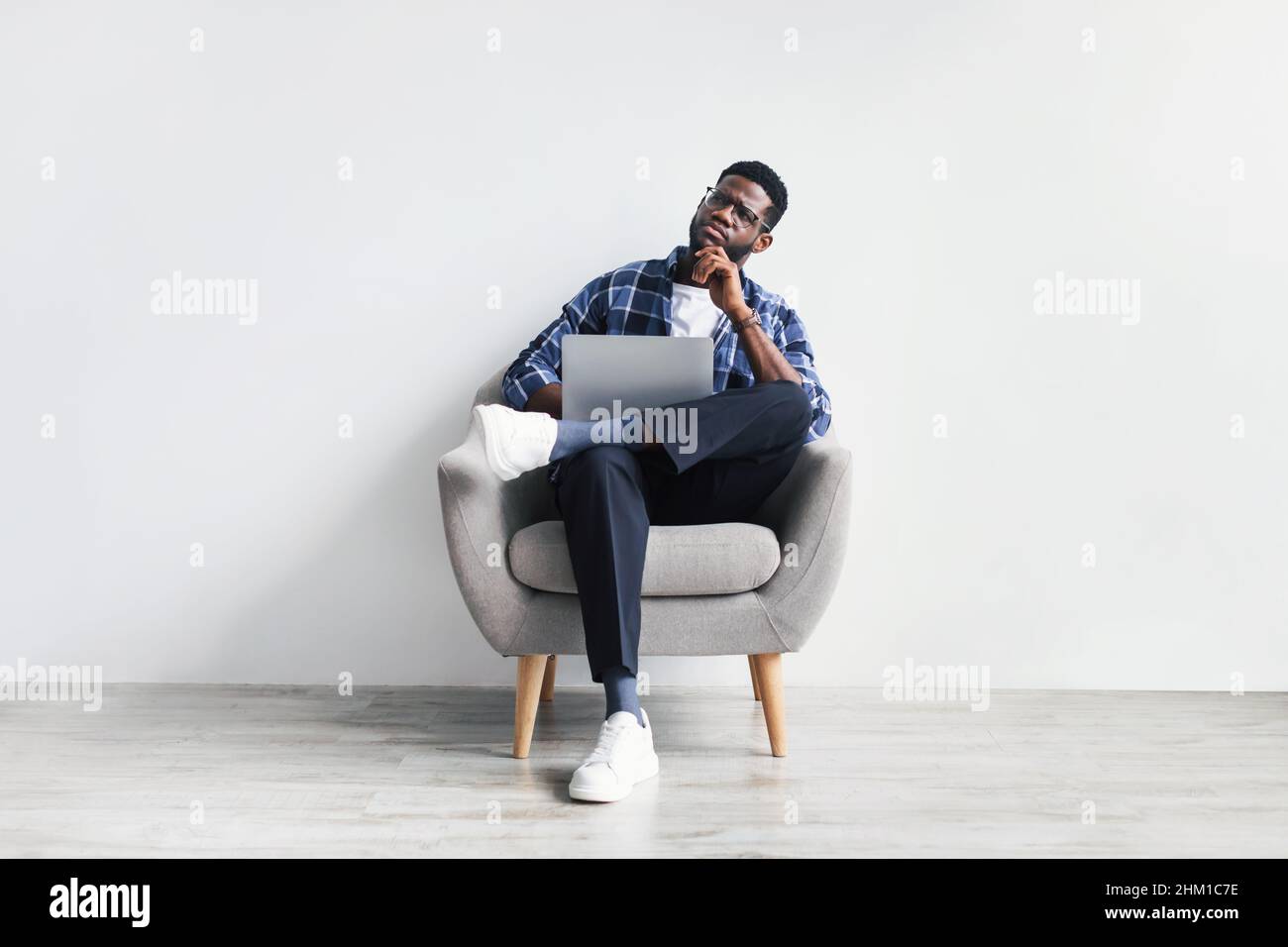Thoughtful young black man using laptop while sitting on chair against ...