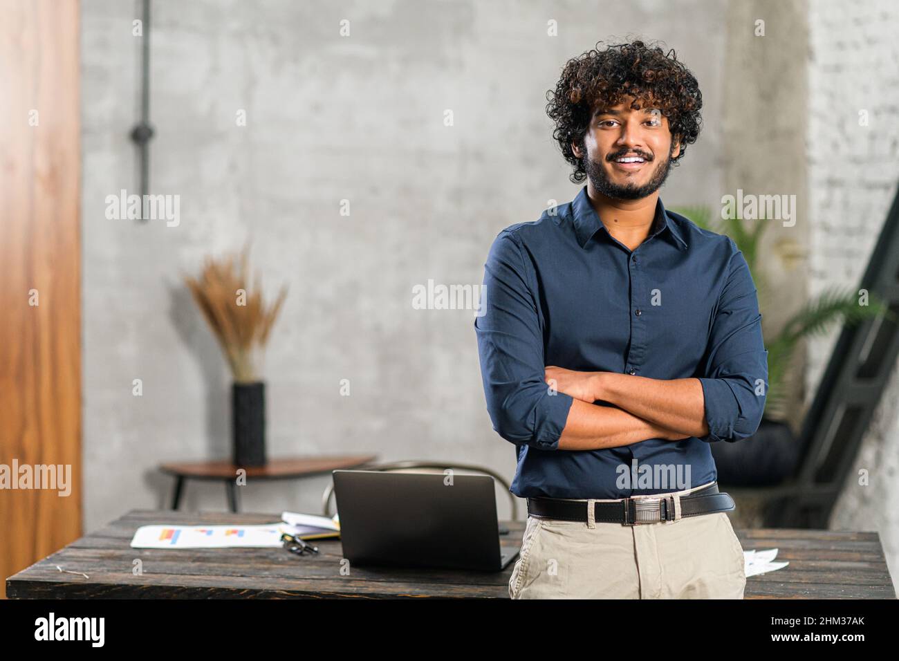 Smiling curly middle eastern businessman standing near desk and looking ...