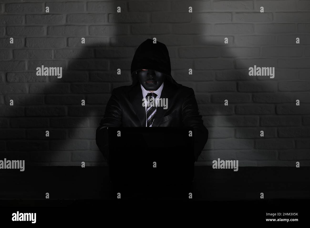 Hacker in black mask and hood at the table in front of the monitor ...