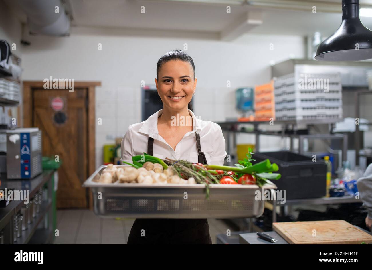 Female chef carrying tray with fresh vegetables and looking at camera ...