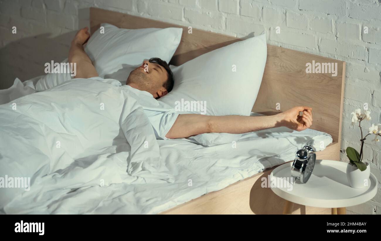 awakened man smiling while stretching in bed near alarm clock on ...