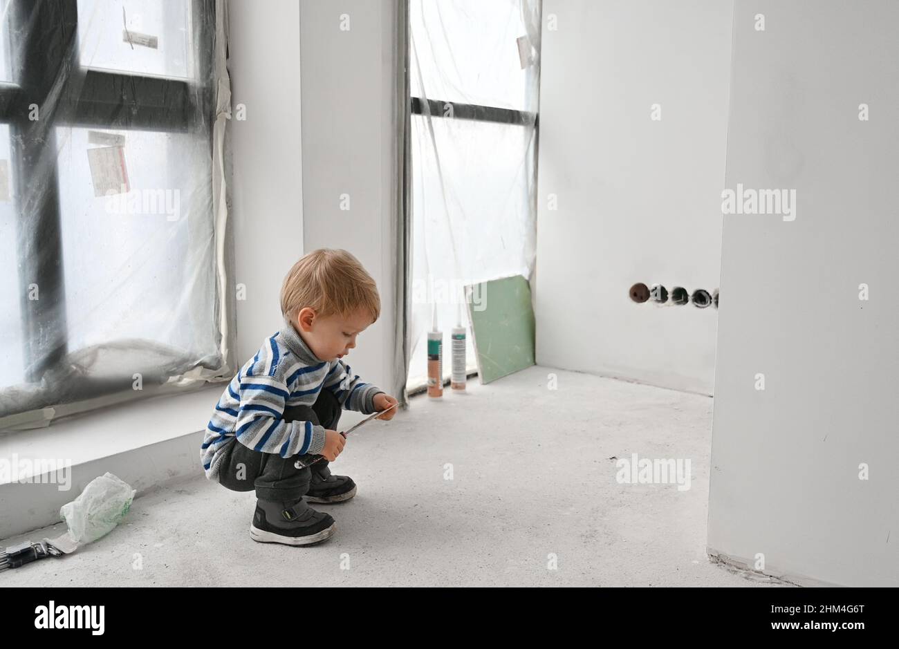 Adorable baby boy crouching down and holding instrument in new home ...