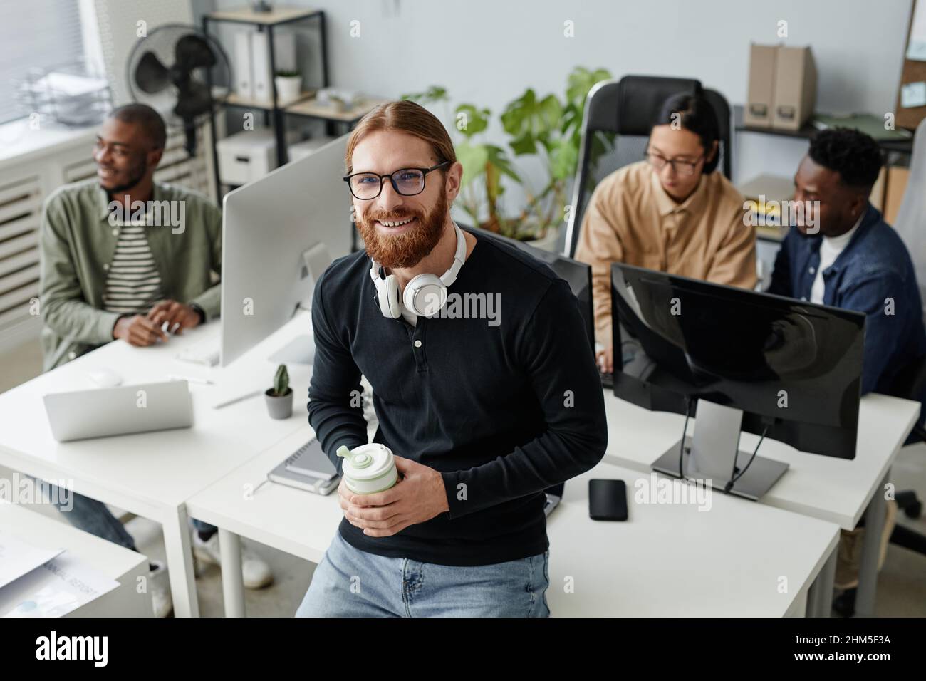 Happy young businessman with drink standing by desk with computer ...