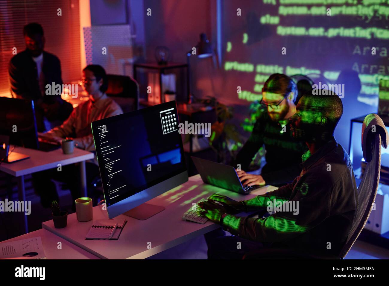 Group of young intercultural office workers sitting by desks in front of computers and decoding ...