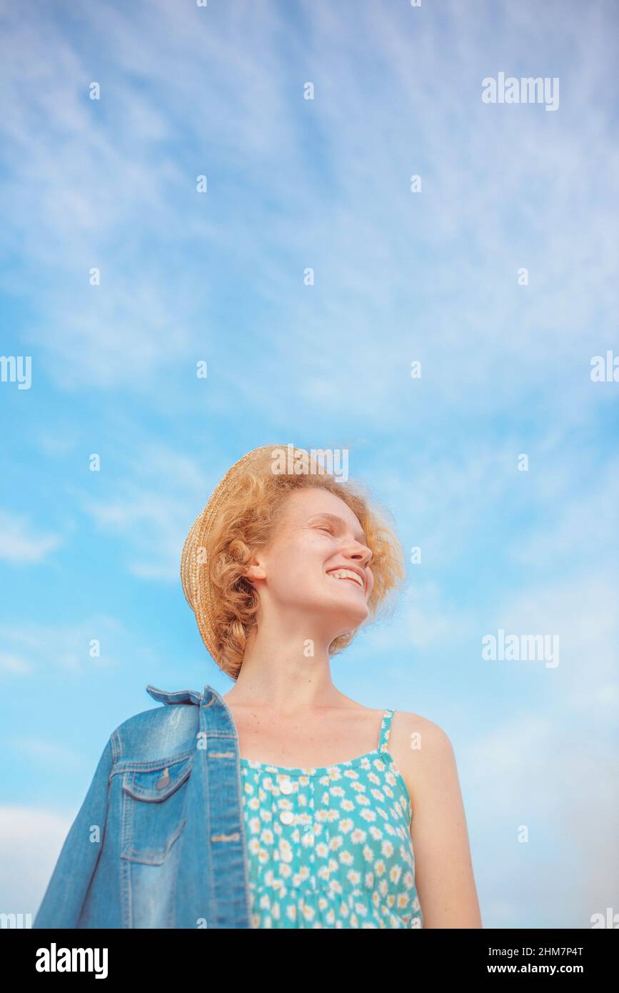 young curly redhead woman in straw hat, blue sundress and jeans jacket ...